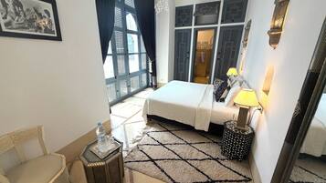 Quarto de hotel com cama, mesa de cabeceira com abajur, cadeira, tapete estampado e uma grande janela com cortinas.