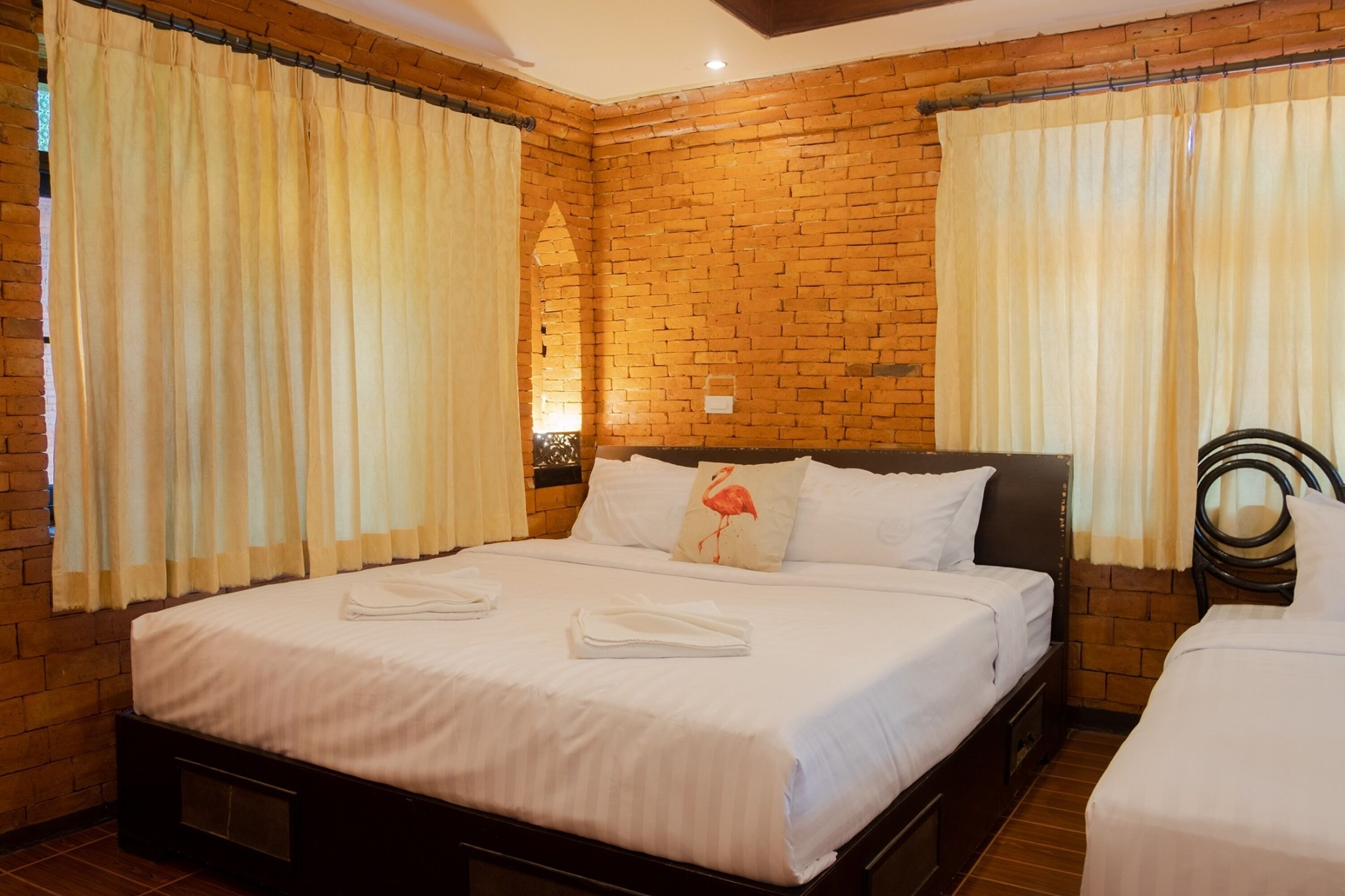 Quarto de hotel com parede de tijolos, uma cama grande com roupa de cama branca, duas mesas de cabeceira e cortinas.