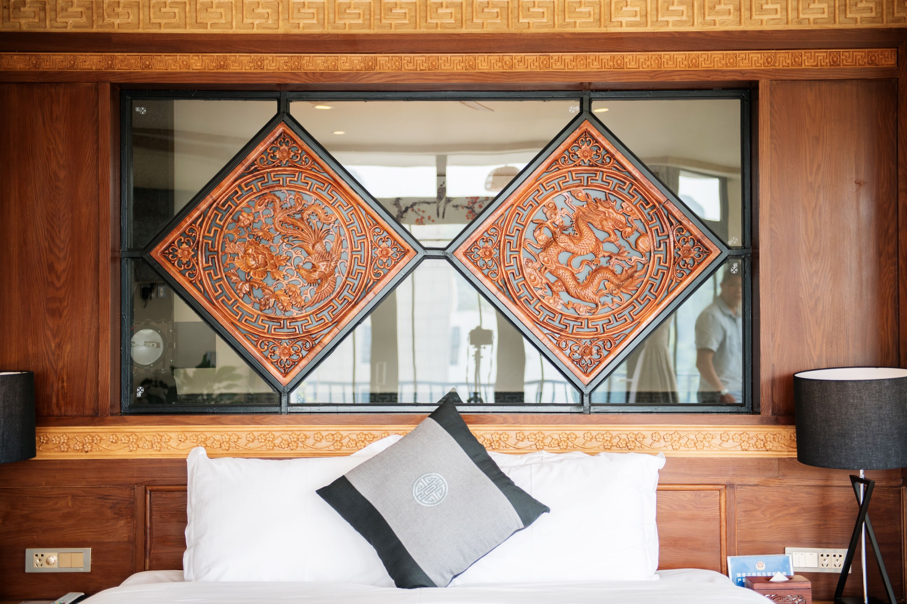 Quarto de hotel com cabeceira de madeira, uma janela grande com desenhos detalhados, cama com roupa de cama branca e almofada cinza, e um abajur preto.