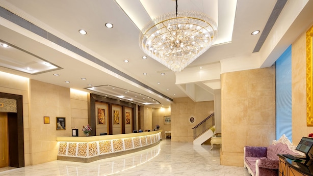 Um saguão de hotel com balcão de recepção, um candelabro, uma escadaria e um sofá roxo.