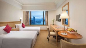 Quarto de hotel com duas camas, uma escrivaninha com cadeira, uma mesa de jantar com uma tigela de frutas, um abajur e uma janela grande com vista para a cidade.
