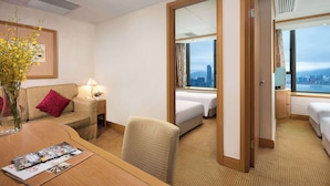 Quarto de hotel com cama, sofá, escrivaninha com revista e vista da cidade através de grandes janelas.