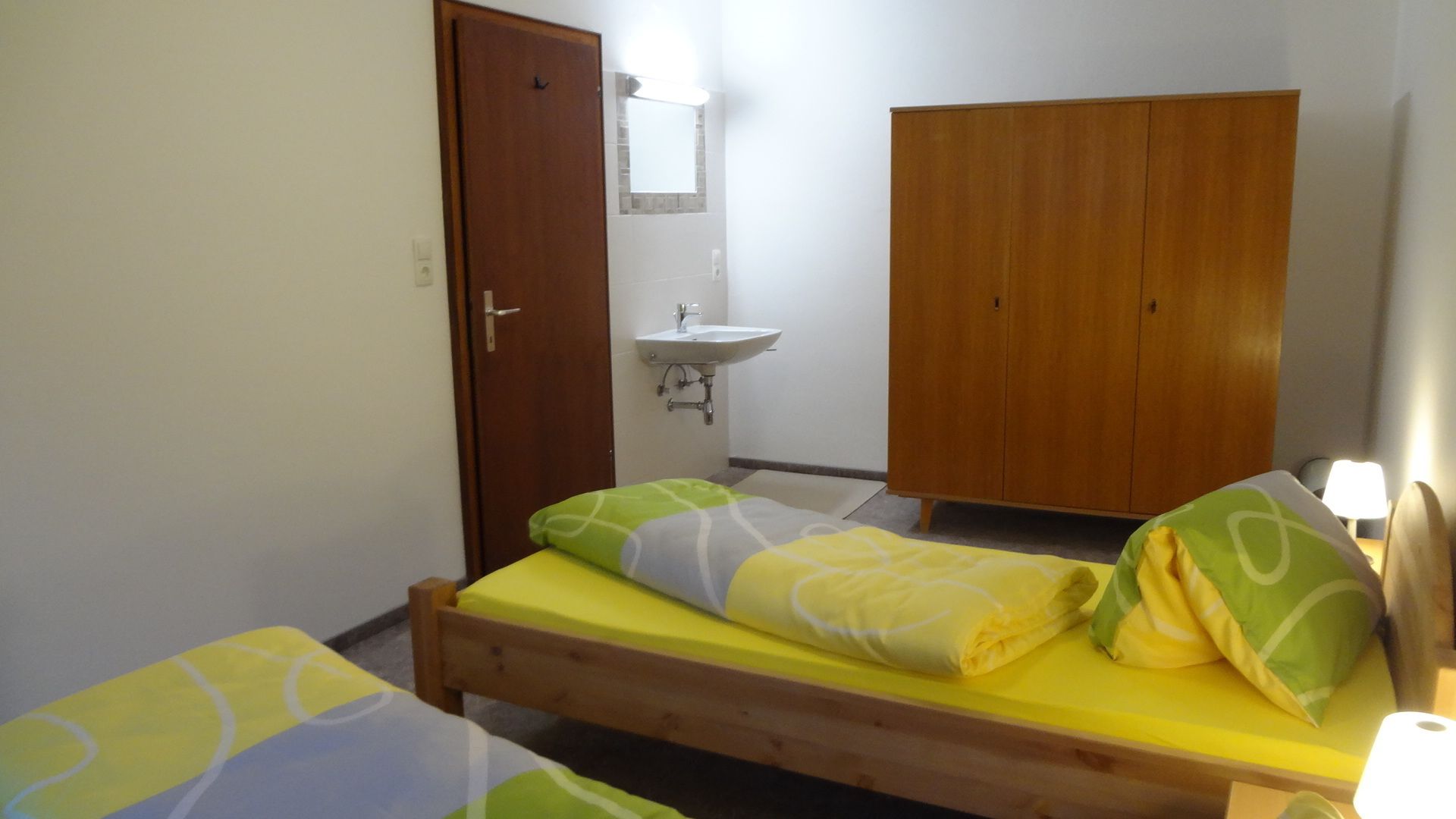 Um quarto pequeno e simples com uma cama, um guarda-roupa de madeira e uma casa de banho com uma pia.