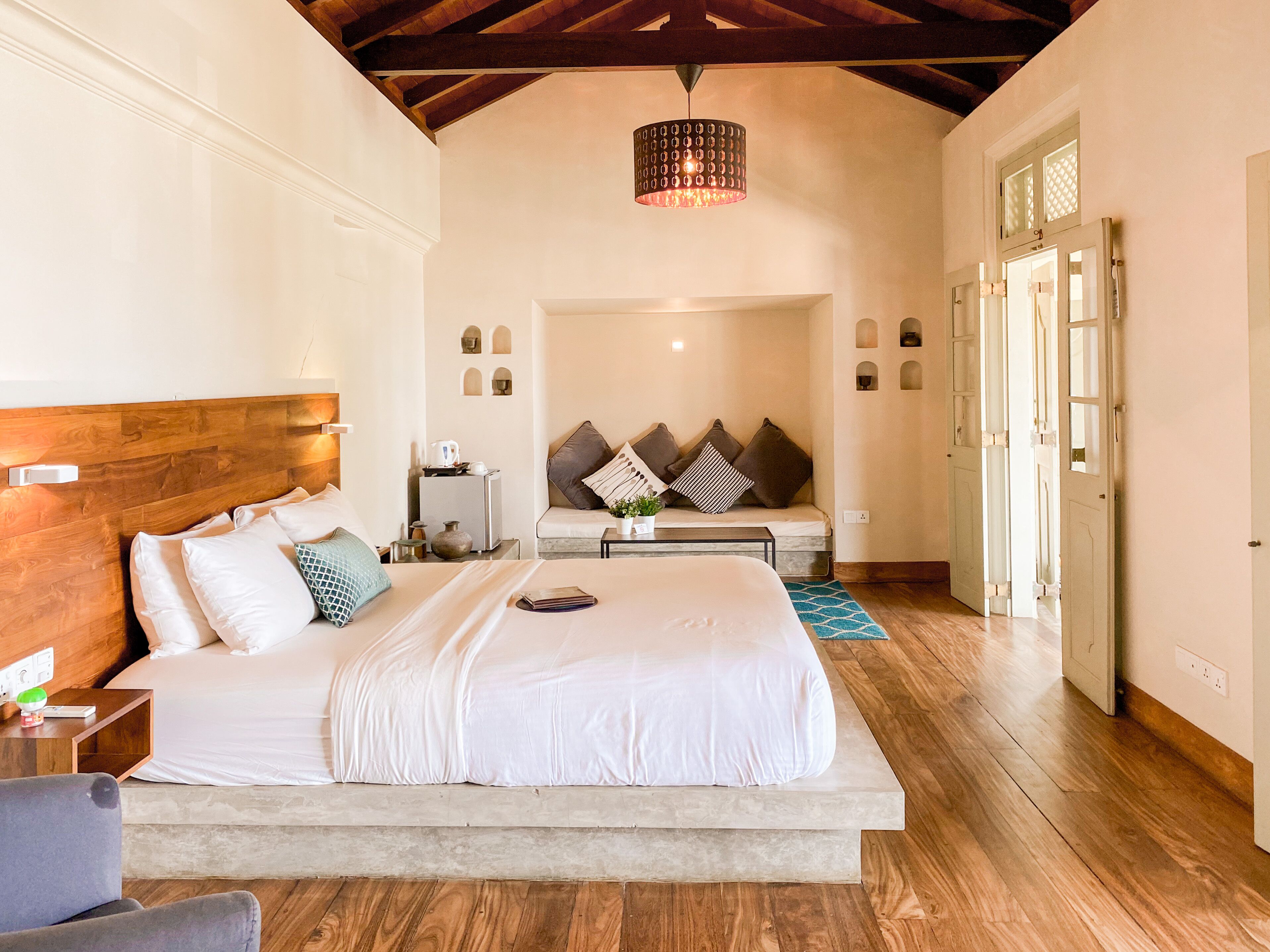 Um quarto espaçoso com uma cama grande, uma cabeceira de madeira, mesa de cabeceira, escrivaninha, cadeira, sofá com almofadas, luminária pendente e uma porta com janela.