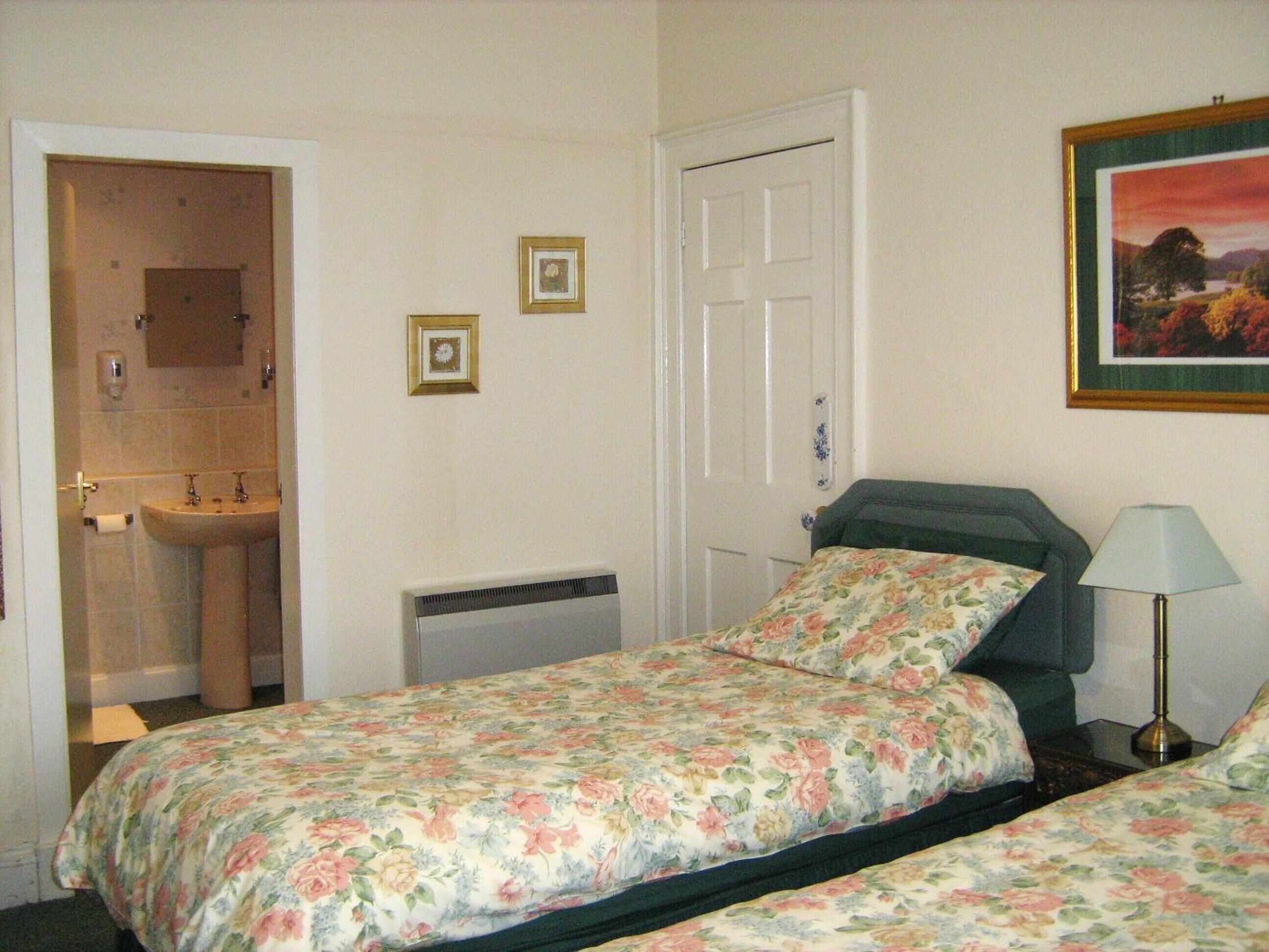 Quarto com duas camas de solteiro, mesa de cabeceira com abajur, porta, banheiro pequeno com pia e um quadro na parede.