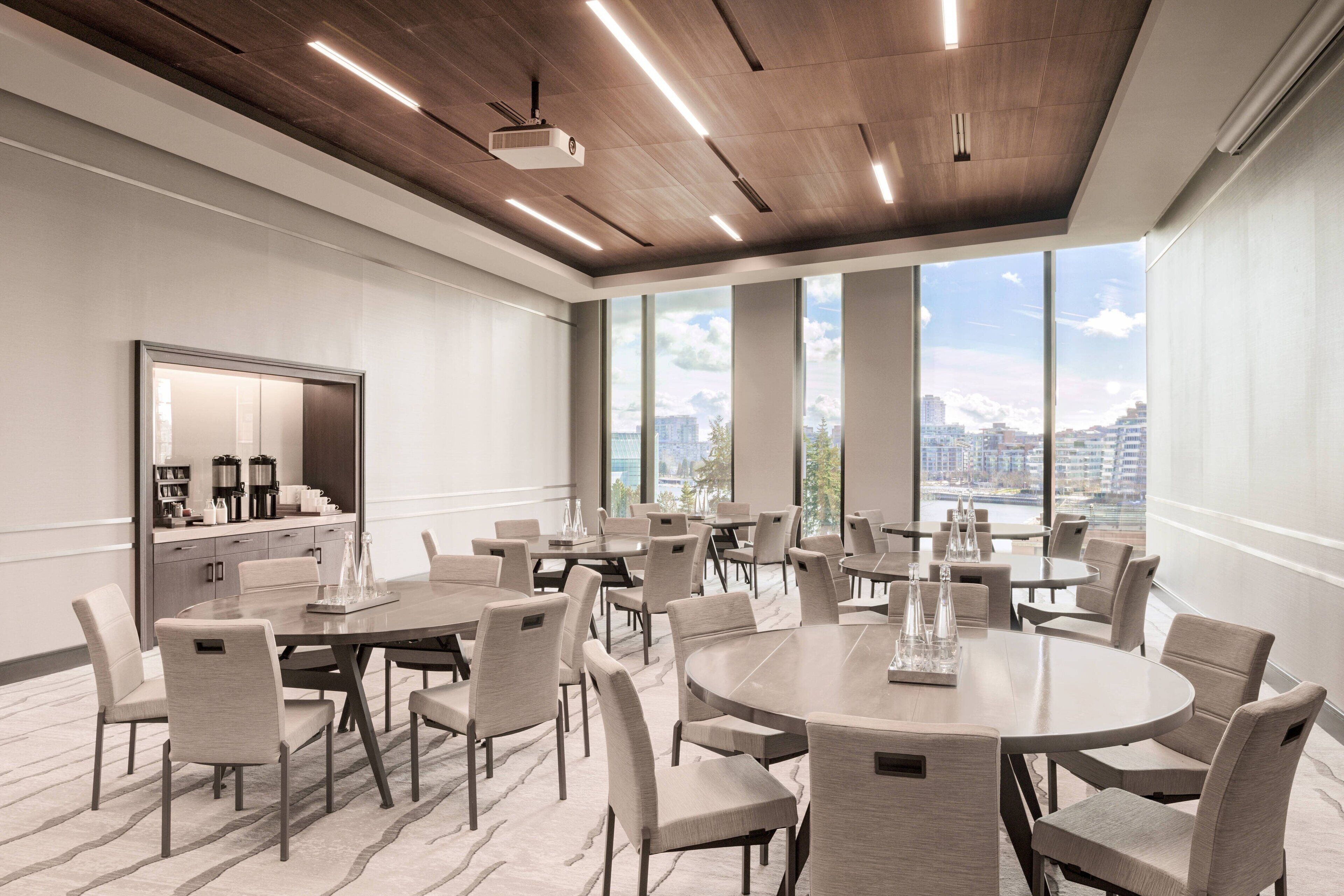 Uma sala de conferências moderna com mesas e cadeiras redondas, grandes janelas com vista para a cidade e um espaço com cozinha.