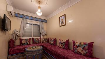 Quarto com sofá, televisão, luminária decorativa no teto e janela com cortinas.