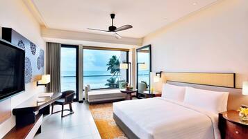 Quarto de hotel com cama, escrivaninha, televisão, ventoinha no teto, janela grande com vista para o oceano e mesa de cabeceira com luminária.