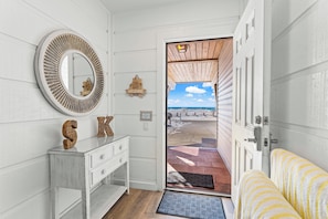 Corredor com porta de madeira que dá para a praia, espelho redondo e letras decorativas em uma cômoda.