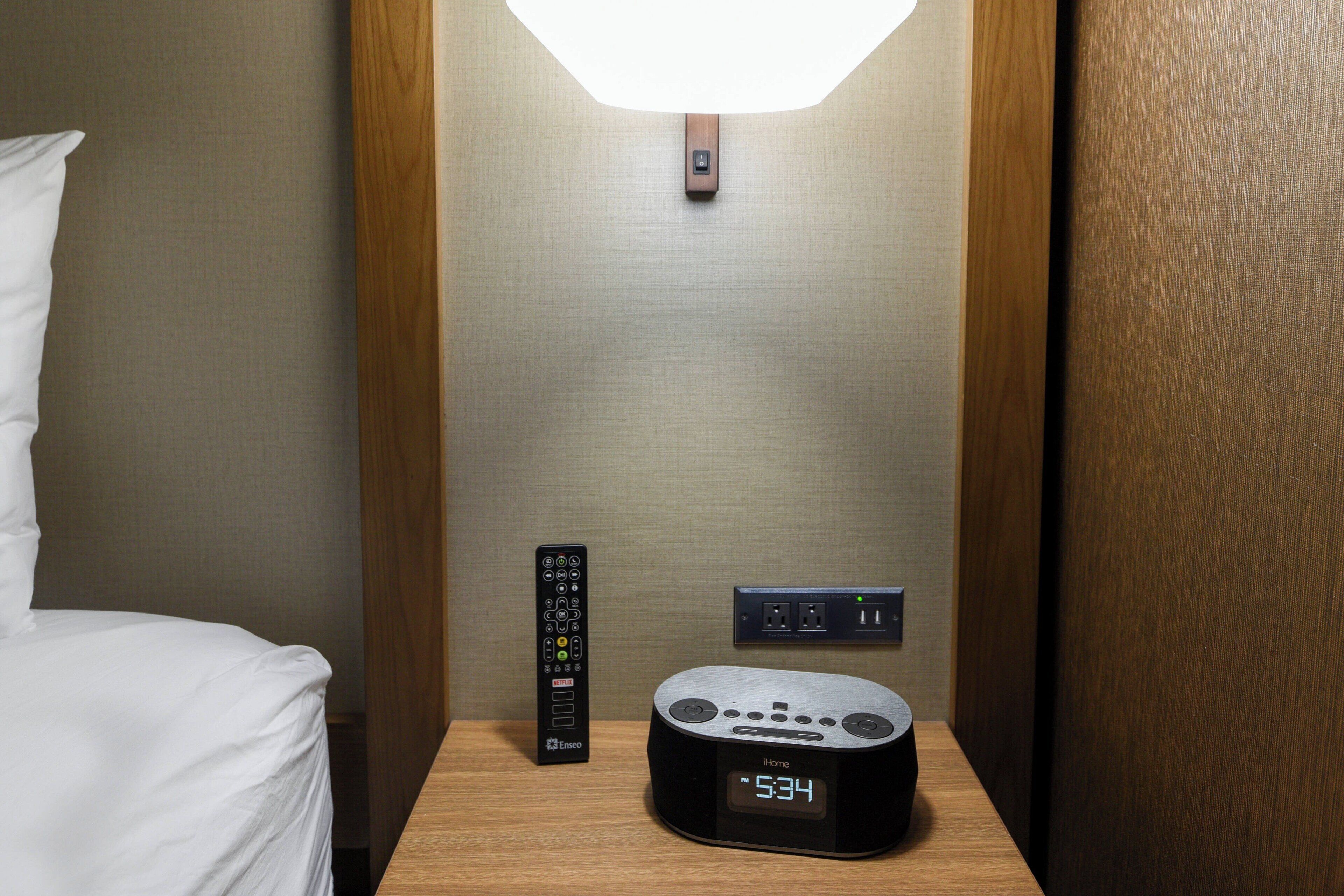 Quarto de hotel com uma cama, mesa de cabeceira, relógio, comando remoto e uma tomada.