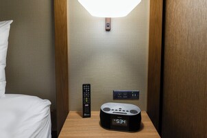Quarto de hotel com uma cama, mesa de cabeceira, relógio, comando remoto e uma tomada.
