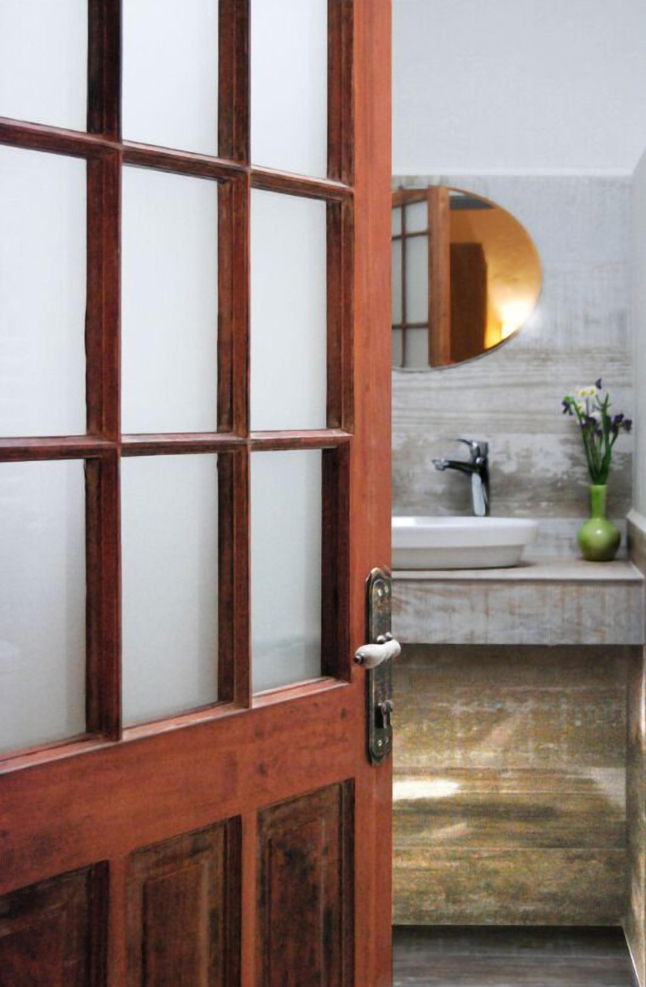 Uma porta de madeira com padrão de grade, que dá para um banheiro com pia, espelho e um vaso com flores.