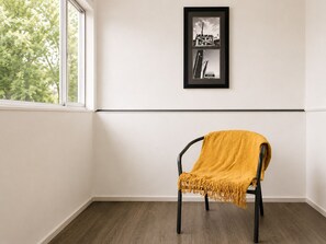 Quarto com uma única cadeira, um quadro na parede e uma janela com vista para árvores.