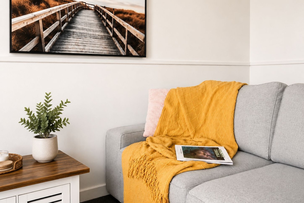 Sala de estar com sofá cinza, manta amarela, uma revista e um quadro com a imagem de uma ponte de madeira.