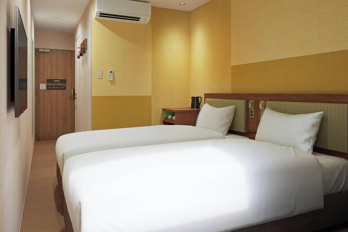 Quarto de hotel com duas camas, porta de madeira, unidade de ar condicionado, mesa de cabeceira e uma pequena mesa com abajur.