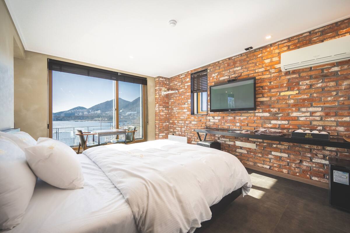 Quarto de hotel moderno com uma cama grande, televisão presa a uma parede de tijolos, varanda com vista para as montanhas e uma pequena mesa com cadeiras.