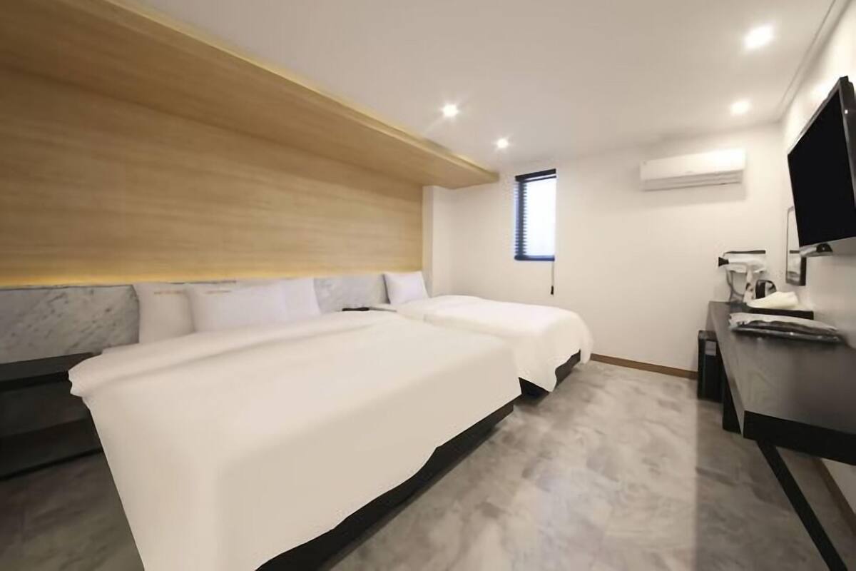 Quarto de hotel moderno com duas camas, uma escrivaninha, televisão de tela plana e janela com persianas.