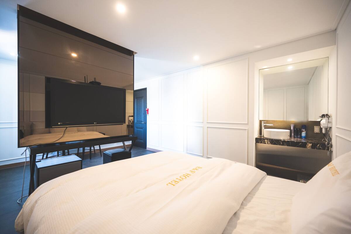 Quarto de hotel moderno com uma cama grande, televisão de tela plana na parede, uma pequena escrivaninha e banheiro com espelho e pia.