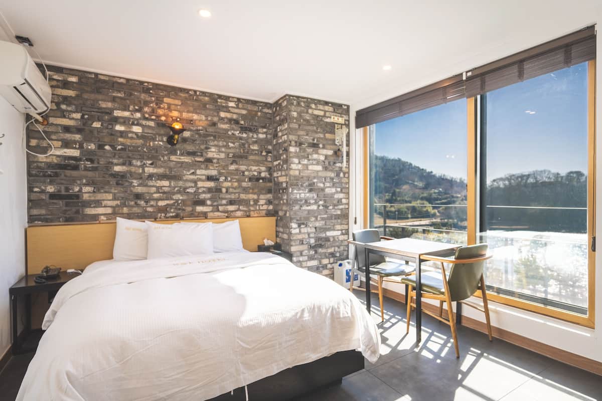 Quarto de hotel moderno com uma cama grande, parede de tijolos decorativa, escrivaninha com cadeira e uma janela ampla com vista para o exterior.