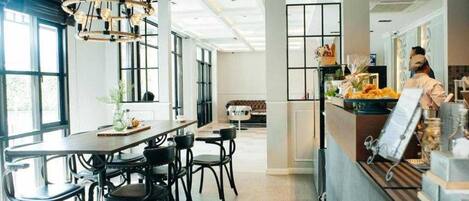 Interior moderno com área de jantar, mesa de madeira e cadeiras pretas, cozinha com bancada exibindo alimentos e grandes janelas que permitem entrada de luz natural.