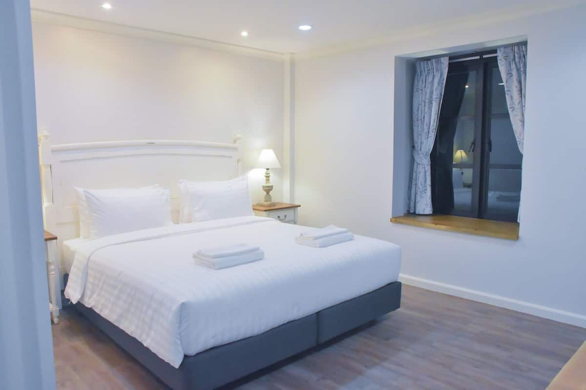 Uma cama bem arrumada com lençóis brancos, uma luminária de cabeceira e uma janela com cortinas.