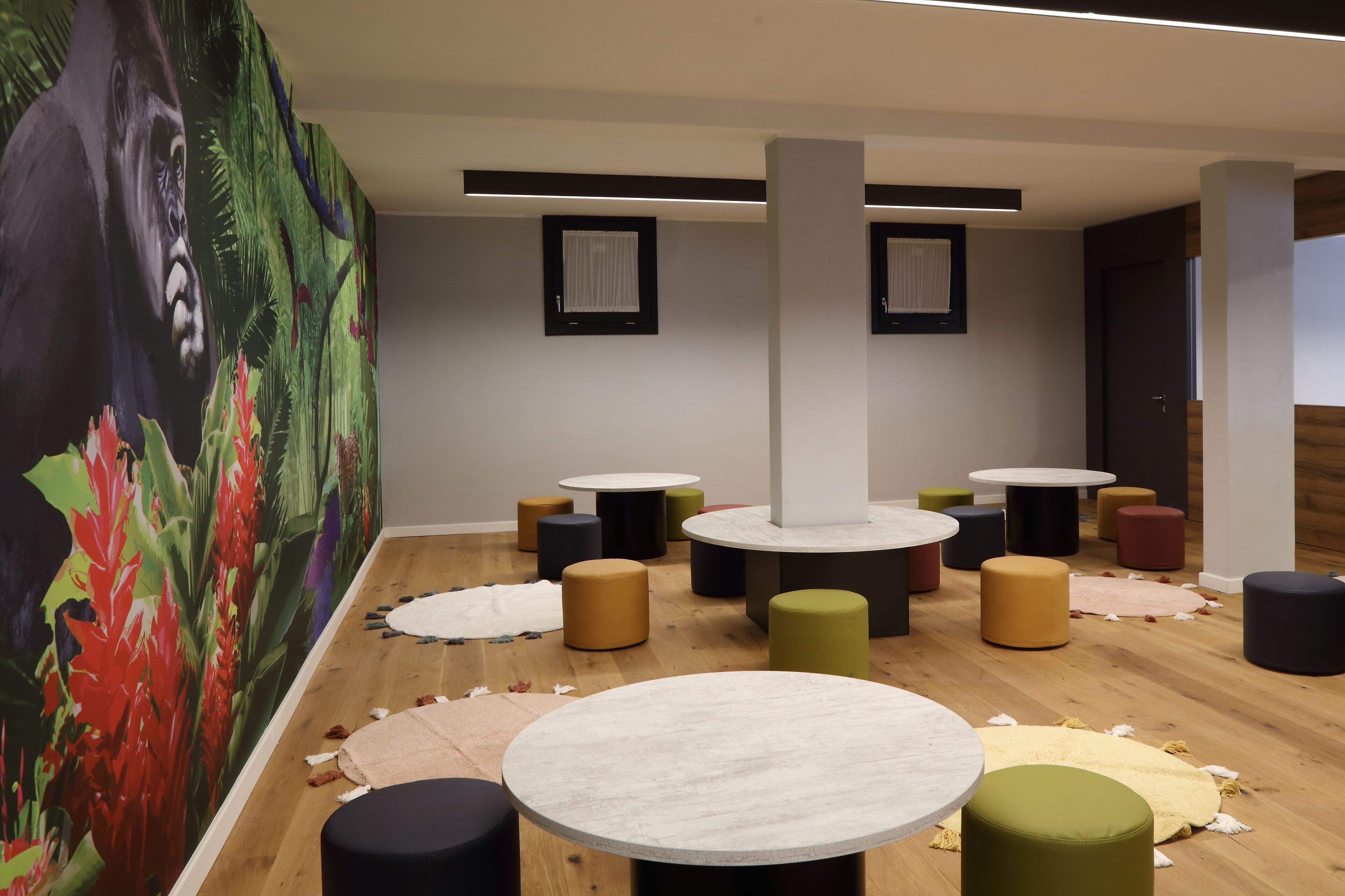 Um interior moderno com mesas redondas e banquetas coloridas, um grande mural e piso de madeira.