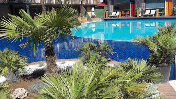 Uma área de piscina com uma piscina azul límpida, cercada por plantas de palmeira e pedrinhas brancas. Prédios de madeira com telhados vermelhos aparecem ao fundo.