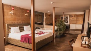 Quarto de hotel com uma cama grande, cabeceira de madeira, uma mesa com chaleira e um vaso de planta.
