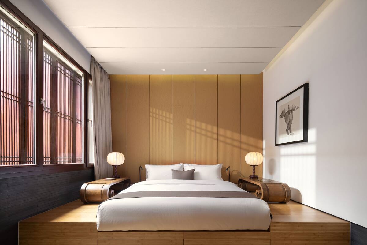 Quarto de hotel moderno com uma cama grande, cabeceira de madeira, duas mesas de cabeceira com luminárias, um quadro na parede e uma janela com persianas verticais.