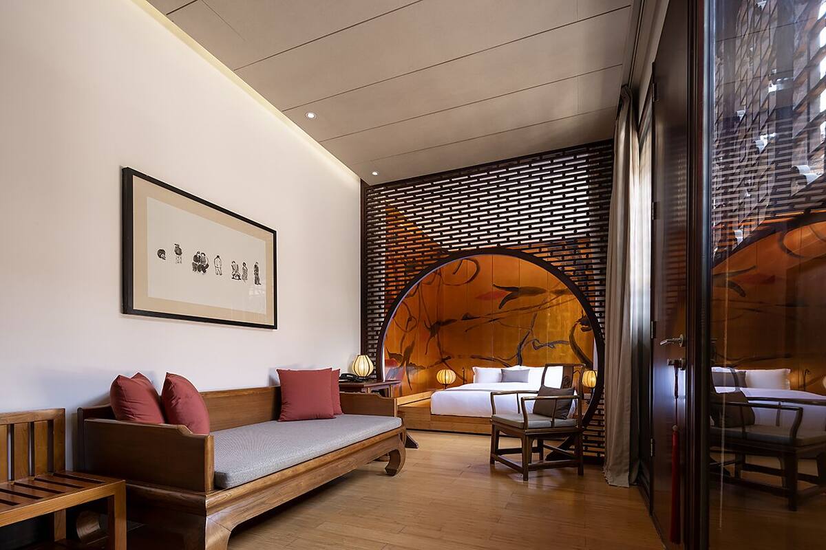 Quarto de hotel moderno com piso de madeira, sofá com almofadas vermelhas, cama com roupa de cama branca e parede com design de treliça de madeira.