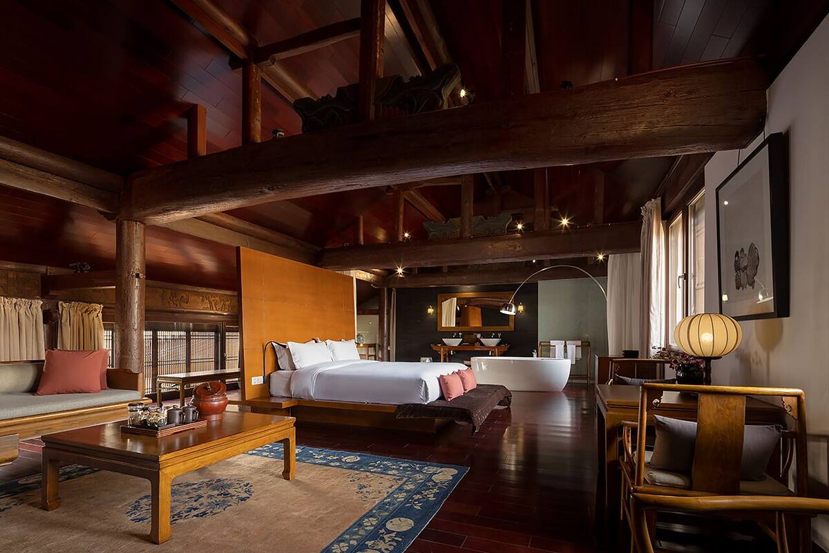 Quarto de hotel espaçoso com uma cama grande, área de estar com sofá e mesa de centro, banheiro com banheira e vigas de madeira no teto.