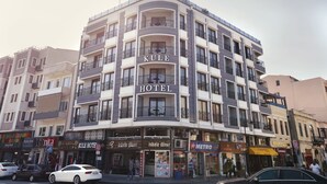 Um hotel com vários andares, varandas, uma placa com a inscrição "KULE HOTEL" e diversas lojas no térreo.