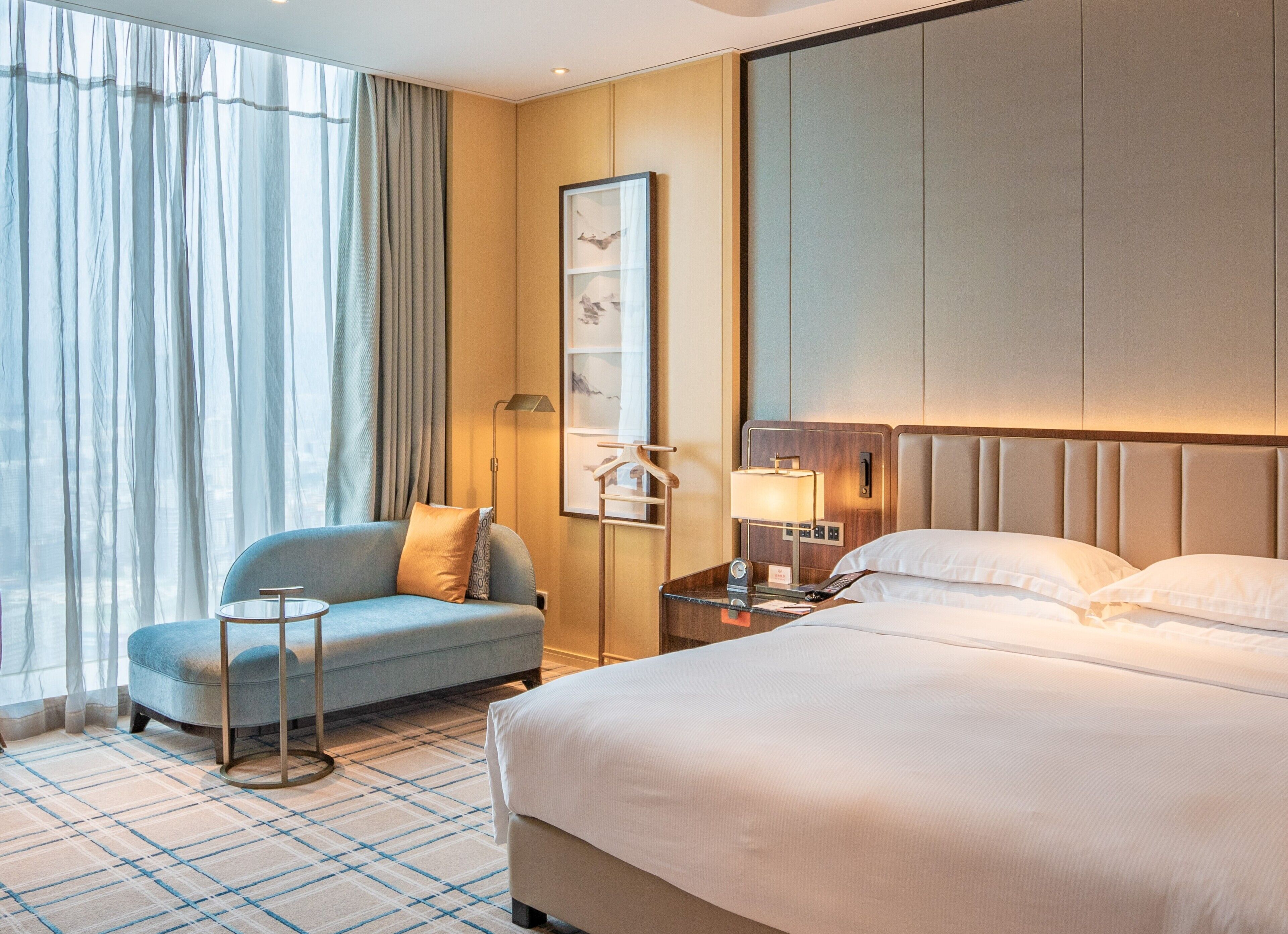Quarto de hotel com cama, uma cadeira, mesa de cabeceira com abajur e uma janela grande com cortinas.