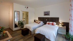 Quarto de hotel com duas camas, piso de madeira, banheiro com chuveiro de vidro e uma pequena mesa com luminária.