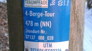 Placa numa árvore com informações sobre a trilha de caminhada 4-Berge-Tour, incluindo a altitude e um número de contato.