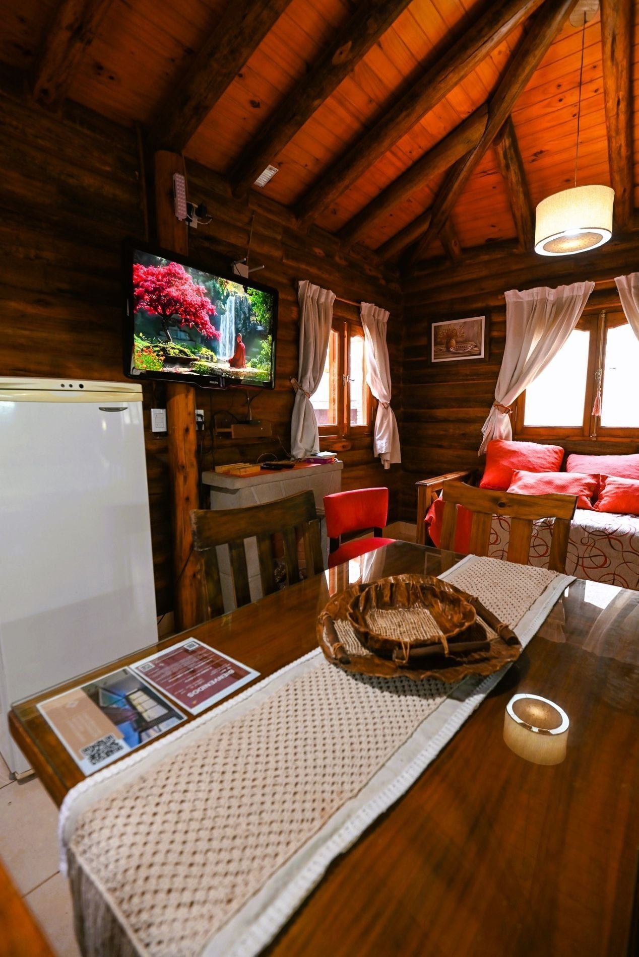Interior de uma cabana de madeira com uma mesa de jantar, cadeiras, uma televisão e um frigorífico.