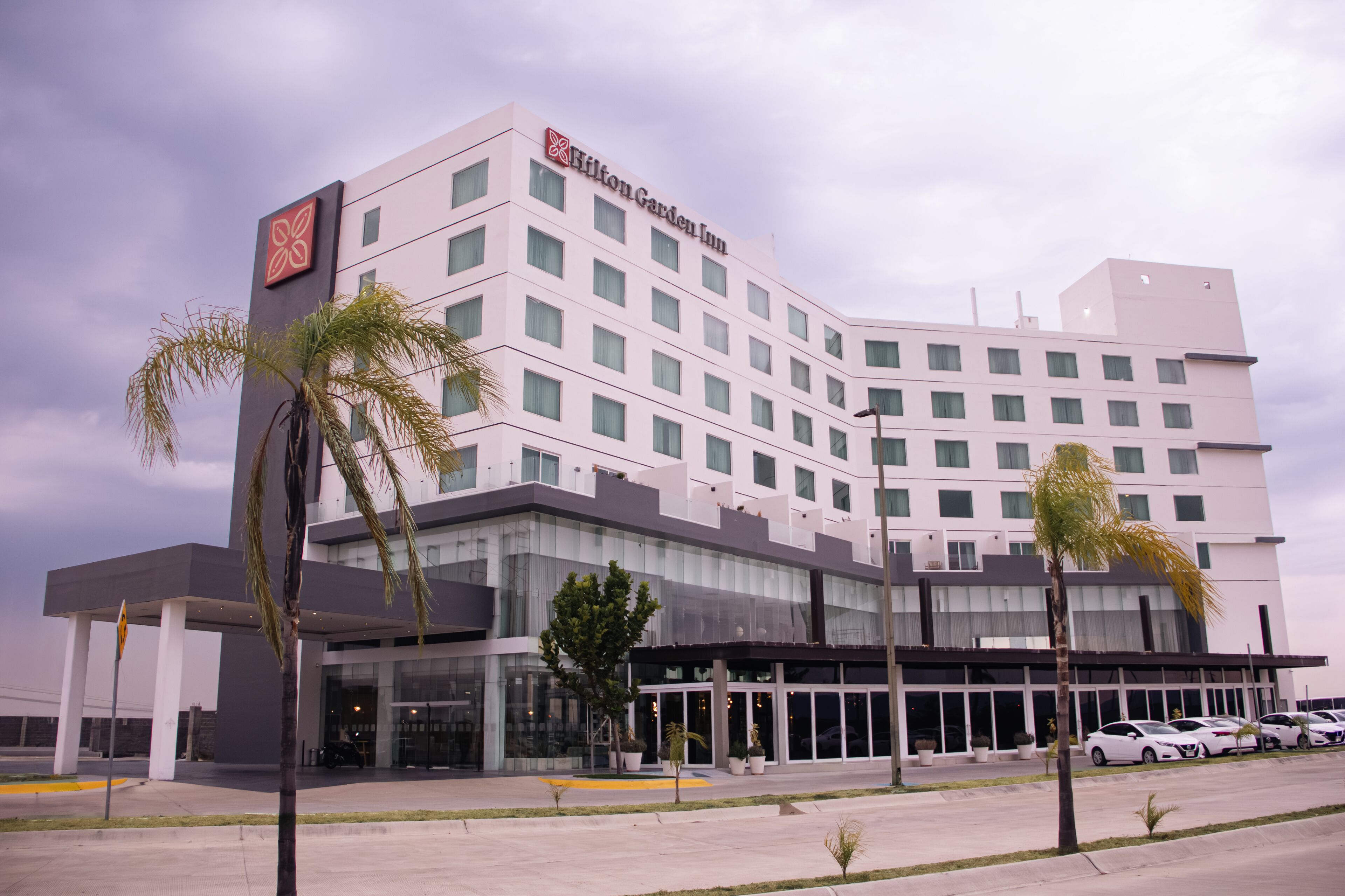 Um Hilton Garden Inn com design moderno, exibindo um logotipo em destaque, vários andares e uma área de estacionamento com diversos carros.