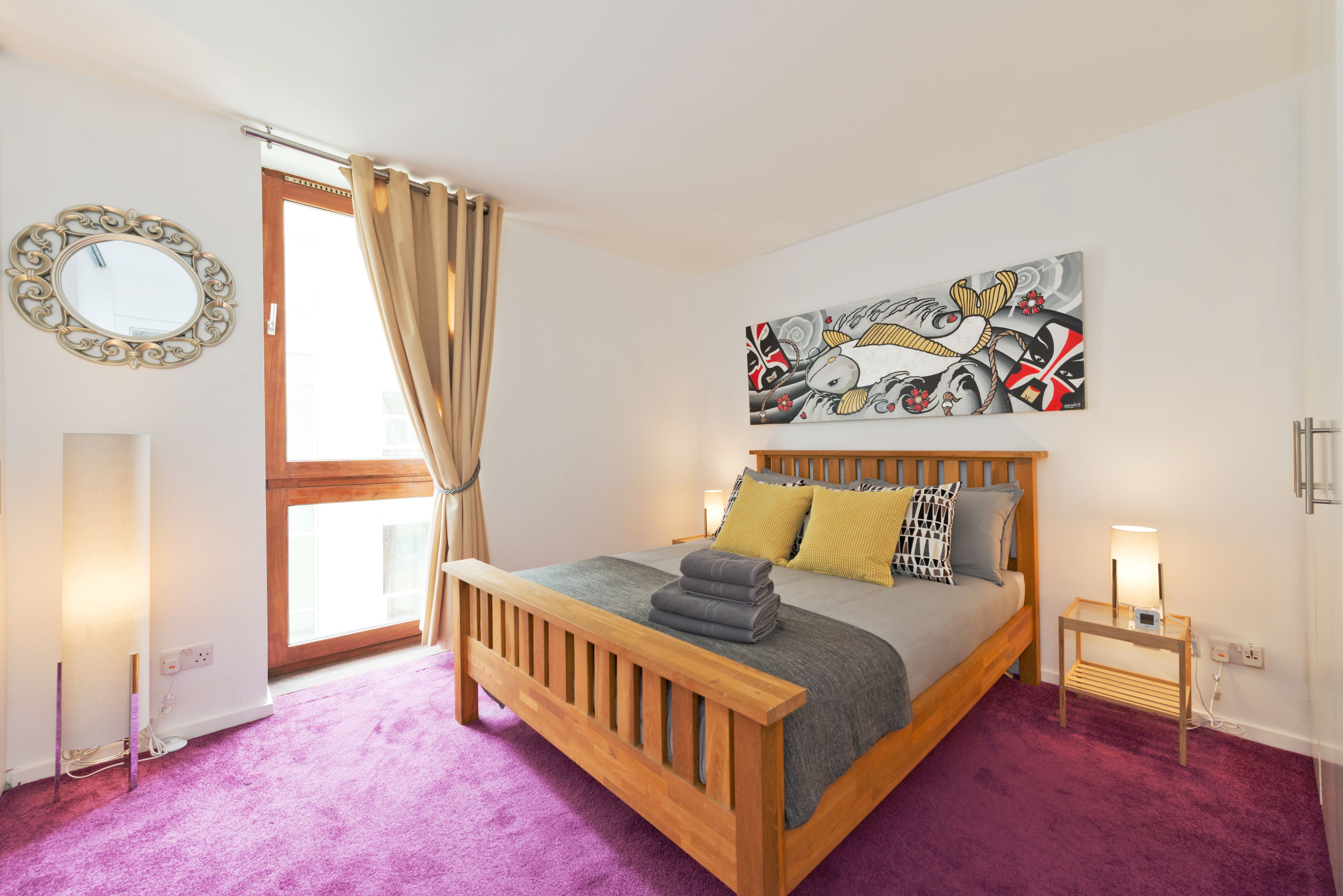 Quarto com cama de madeira, janela com cortinas, espelho fixado na parede e uma obra de arte abstrata.
