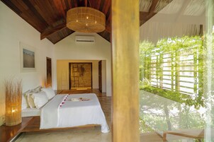 Quarto com cama, teto de madeira, luminária pendente e vista para o verde lá fora.