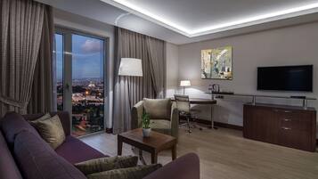 Quarto de hotel moderno com um sofá grande, uma mesinha com uma planta, uma escrivaninha com uma cadeira, uma televisão e vista para a cidade pela janela.