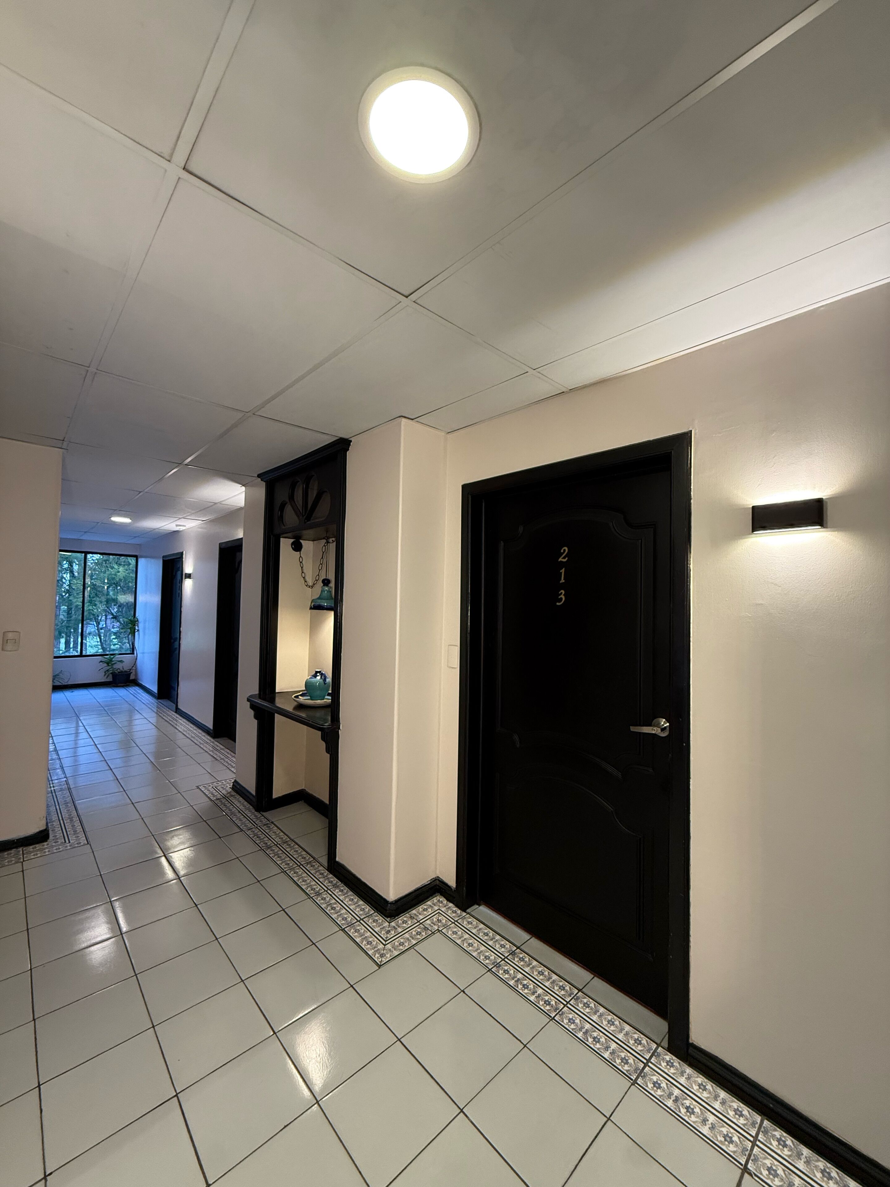 Um corredor de hotel com chão ladrilhado, uma porta preta com número e um acessório de iluminação no teto.