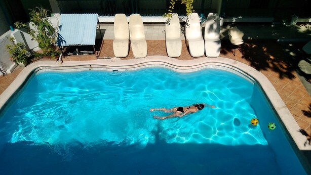 Piscina com uma pessoa nadando, cadeiras de descanso e um deck revestido de azulejos.