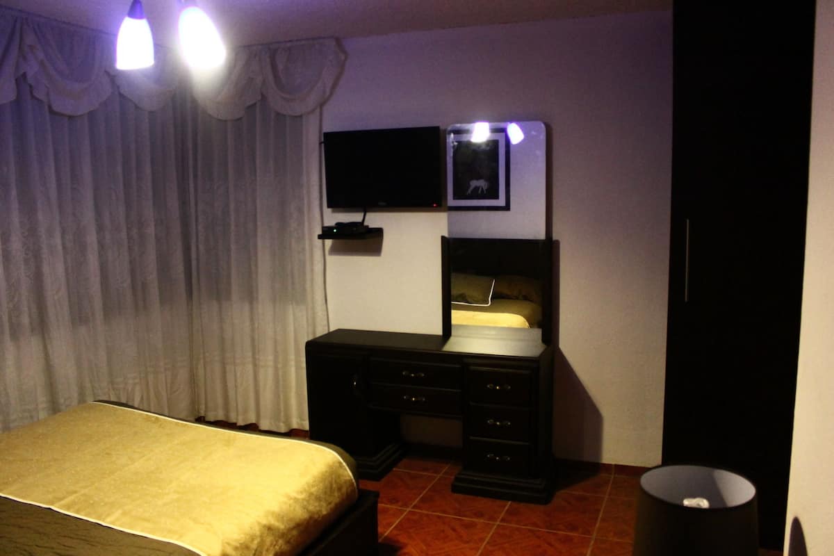 Quarto de hotel com cama, penteadeira, televisão fixada na parede e mesa de cabeceira.