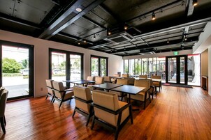 Um espaço amplo e interno com piso de madeira, várias mesas e cadeiras, janelas grandes e um design moderno de teto industrial.
