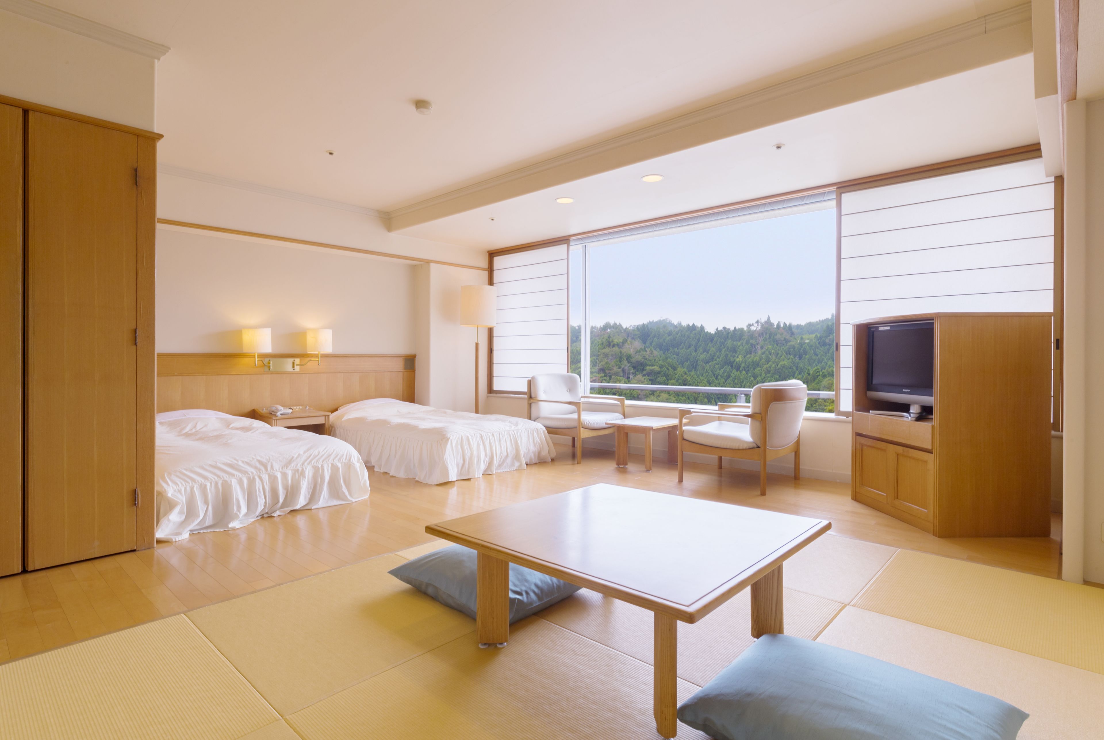 Quarto de hotel moderno com duas camas, televisão, mesa de jantar e vista para a floresta.