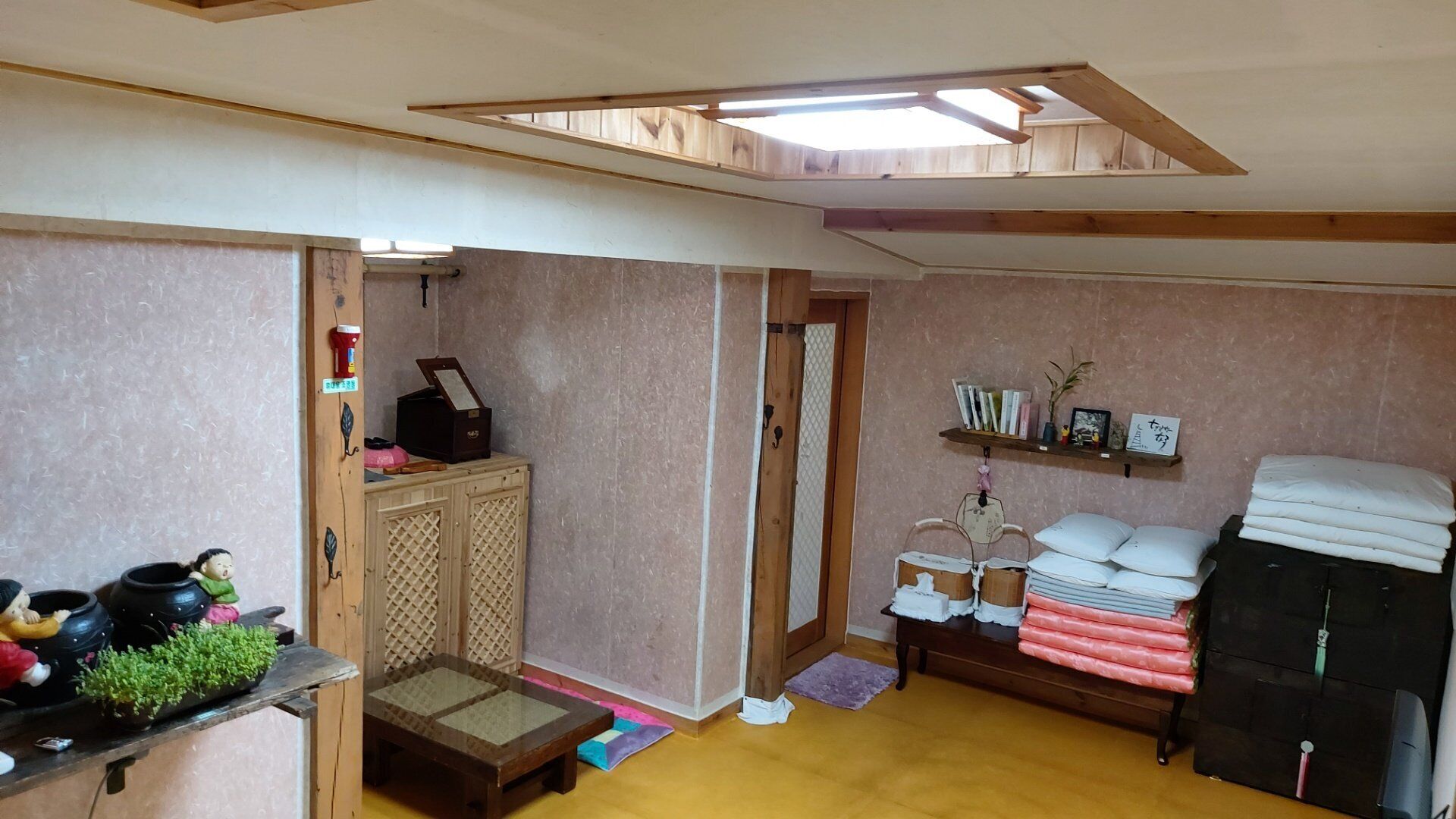 Quarto com teto inclinado, um armário de madeira, uma mesa pequena, uma prateleira com livros e objetos decorativos, e uma cama com almofadas.