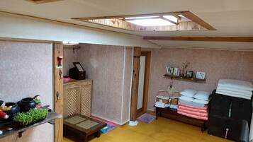 Quarto com teto inclinado, um armário de madeira, uma mesa pequena, uma prateleira com livros e objetos decorativos, e uma cama com almofadas.