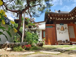 Um edifício em estilo japonês tradicional, com porta de madeira, caminho de pedra e um pequeno jardim com flores vermelhas e uma árvore.
