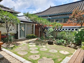 Um pátio tradicional coreano com telhado de telhas, caminhos de pedra e diversas plantas.