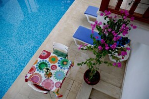 Uma área ao lado da piscina com uma mesa coberta por uma toalha de mesa florida e colorida, um vaso com flores rosa e duas cadeiras de descanso com almofadas azuis.
